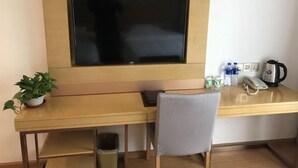 Quarto de hotel com escrivaninha de madeira, televisão fixada acima, uma cadeira, lixeira, planta em vaso e cafeteira.