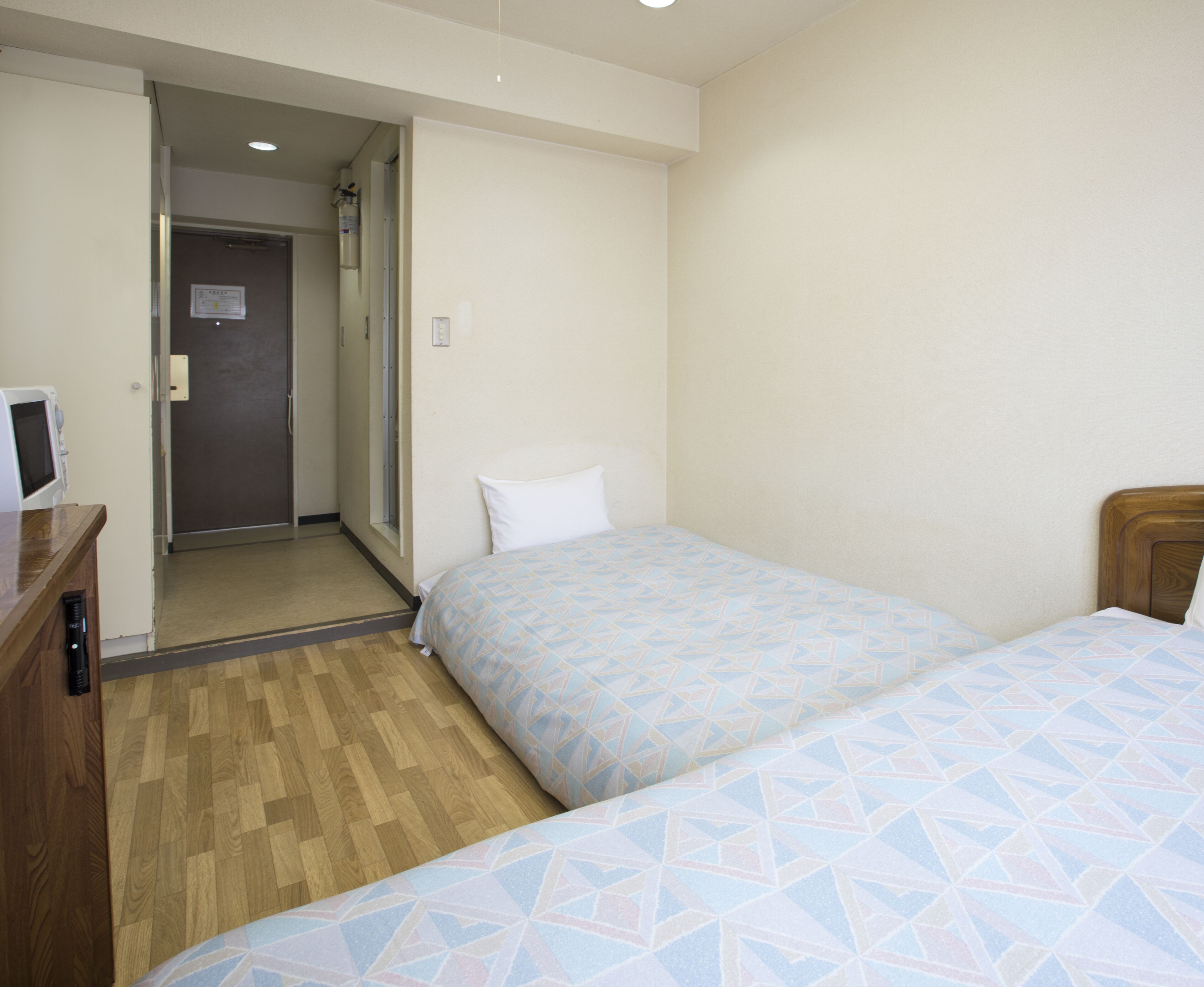 Quarto de hotel com duas camas, uma cômoda de madeira, micro-ondas e porta para um corredor.