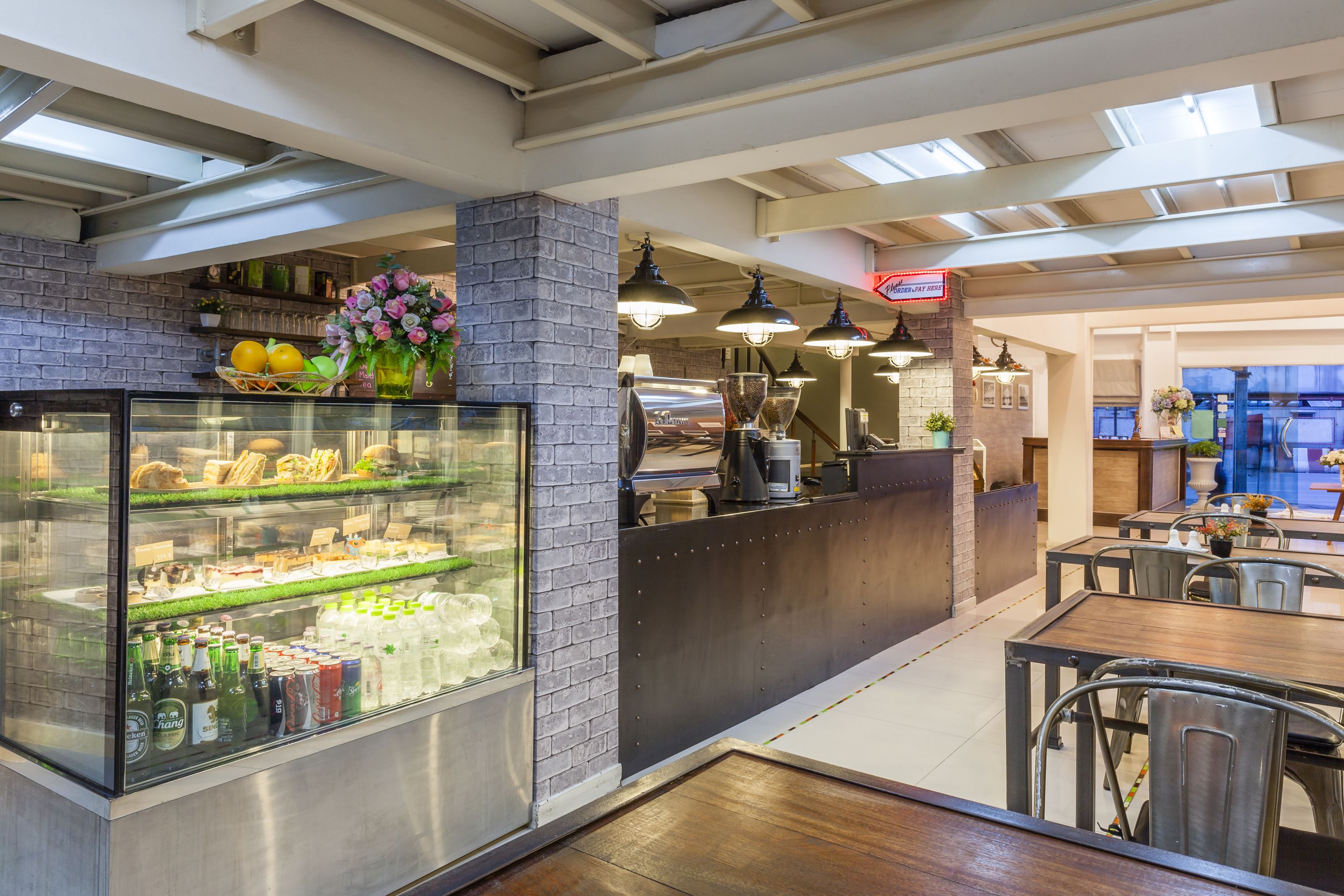 Um restaurante com parede de tijolos, uma vitrine de vidro com comida e bebidas, e uma área de estar com cadeiras e mesas de metal.