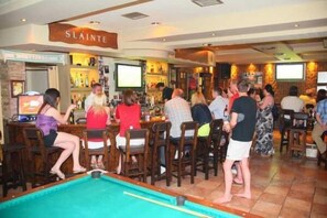 Uma área de bar com mesa de bilhar, cadeiras de madeira e uma placa com a inscrição 'SLAINE'.
