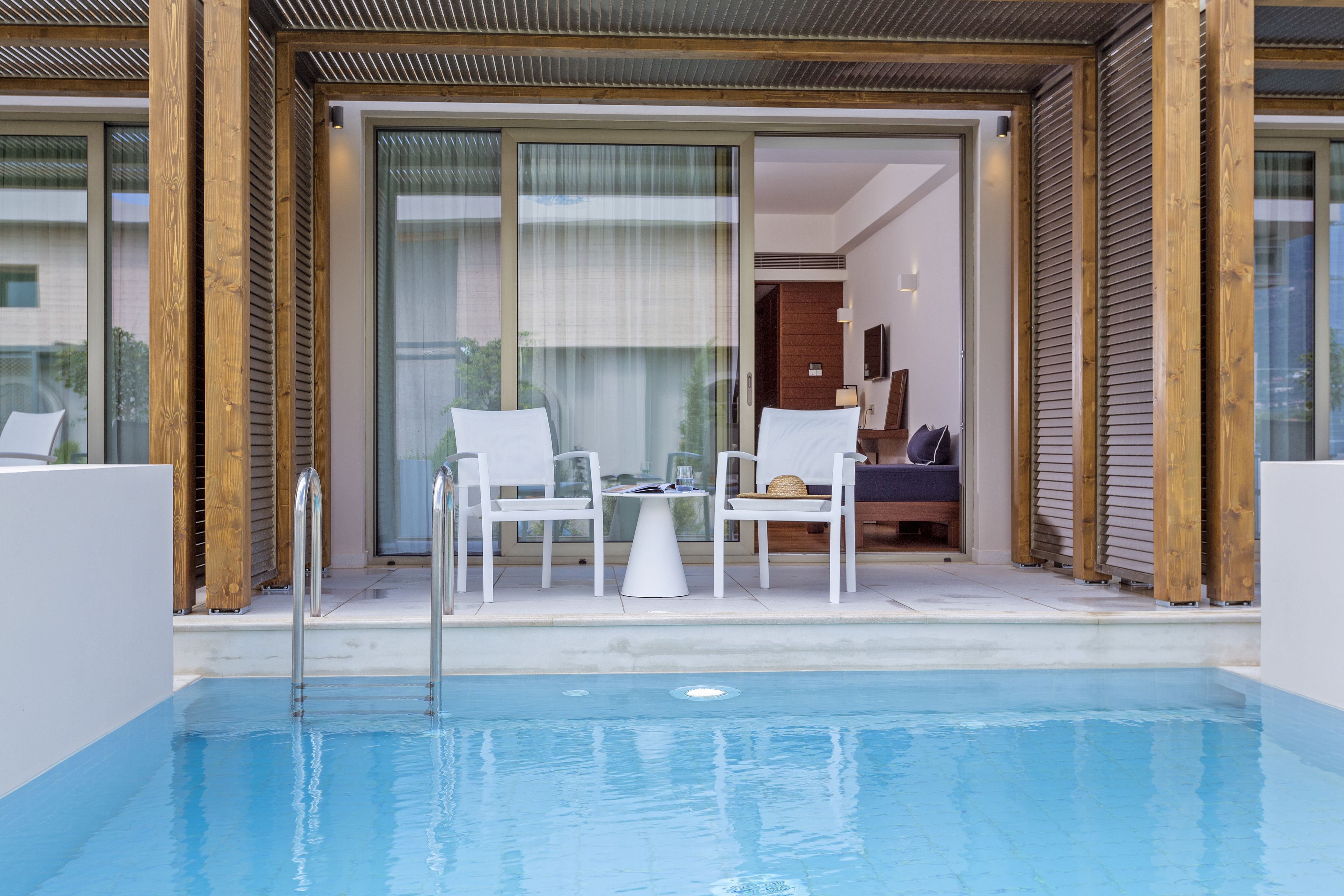Uma área de piscina moderna com duas espreguiçadeiras brancas, uma mesinha e um quarto com paredes de vidro, contendo uma cama e uma televisão.