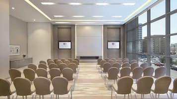 Uma sala de conferências com fileiras de cadeiras voltadas para uma tela de apresentação, grandes janelas com vista para a cidade e piso coberto de carpete.
