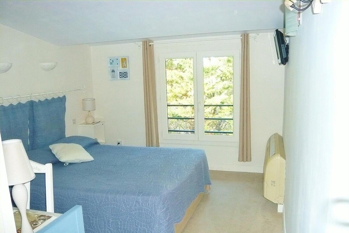 Quarto com cama, mesa de cabeceira, luminária, janela com cortinas e vista para o verde lá fora.