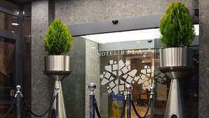 Entrada de hotel com fachada de vidro moderna, duas árvores topiárias em vasos e uma barreira de metal com corda.