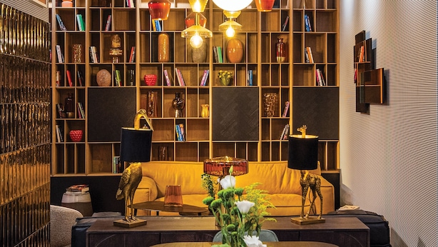 Uma sala de estar acolhedora com um sofá, mesa de centro e luminárias decorativas. Uma estante cheia de livros e objetos decorativos fica ao fundo.
