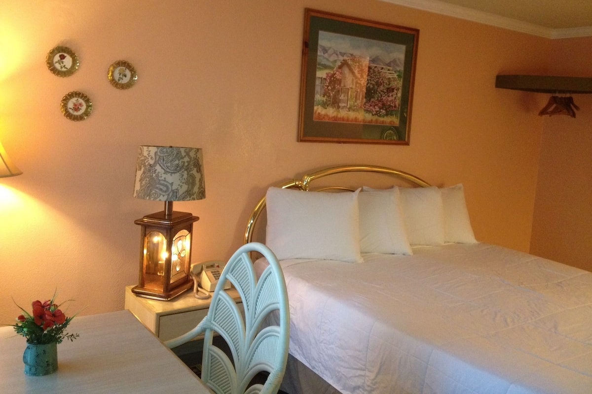 Quarto de hotel com cama, mesa de cabeceira com abajur, uma cadeira e uma pequena mesa com um vaso de flores.