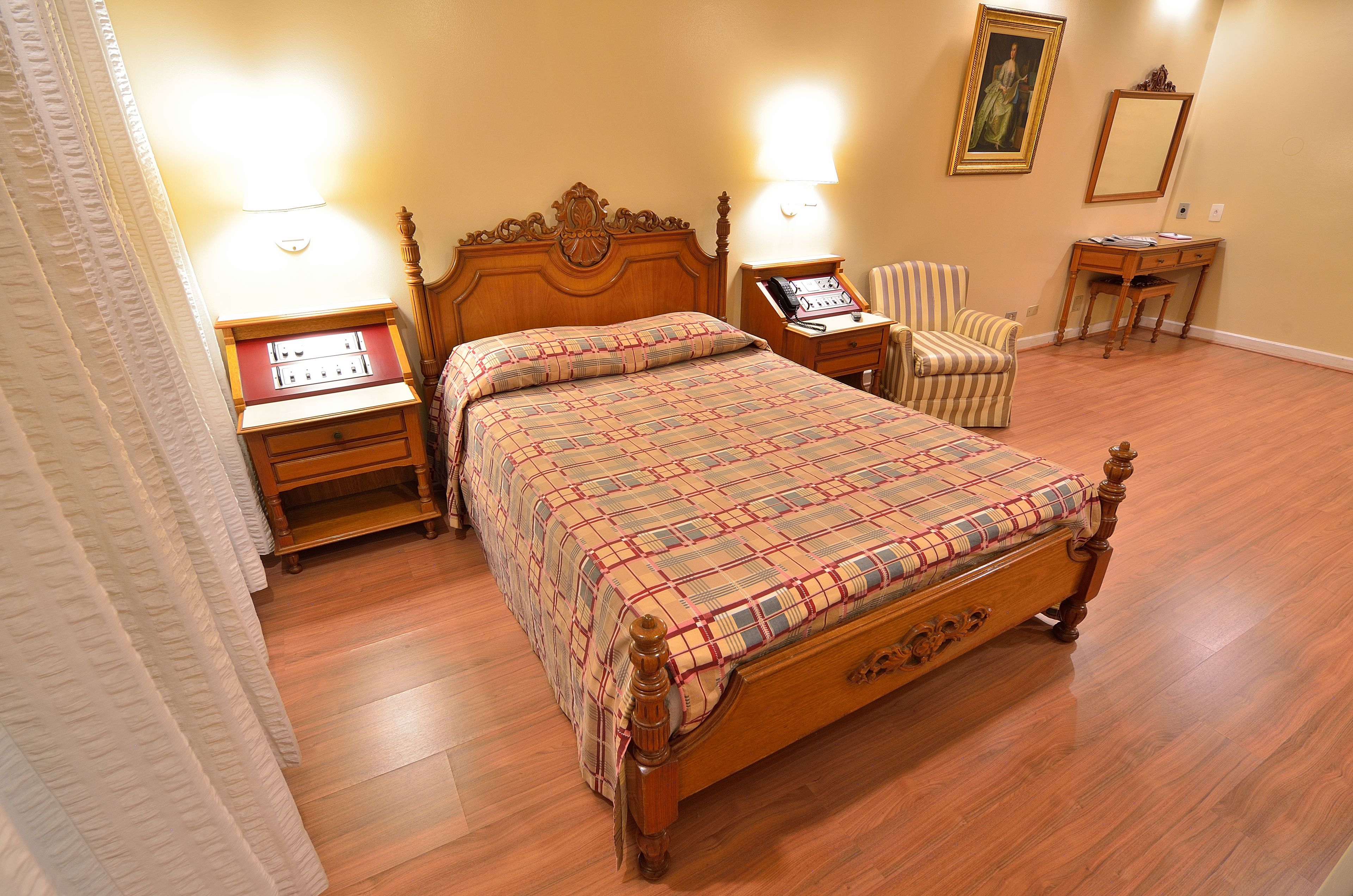 Quarto bem arrumado com cama de madeira, mesas de cabeceira, uma cadeira e uma escrivaninha.