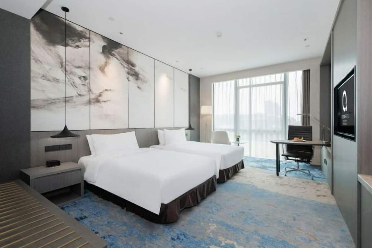 Quarto de hotel moderno com duas camas, uma escrivaninha e uma janela grande.