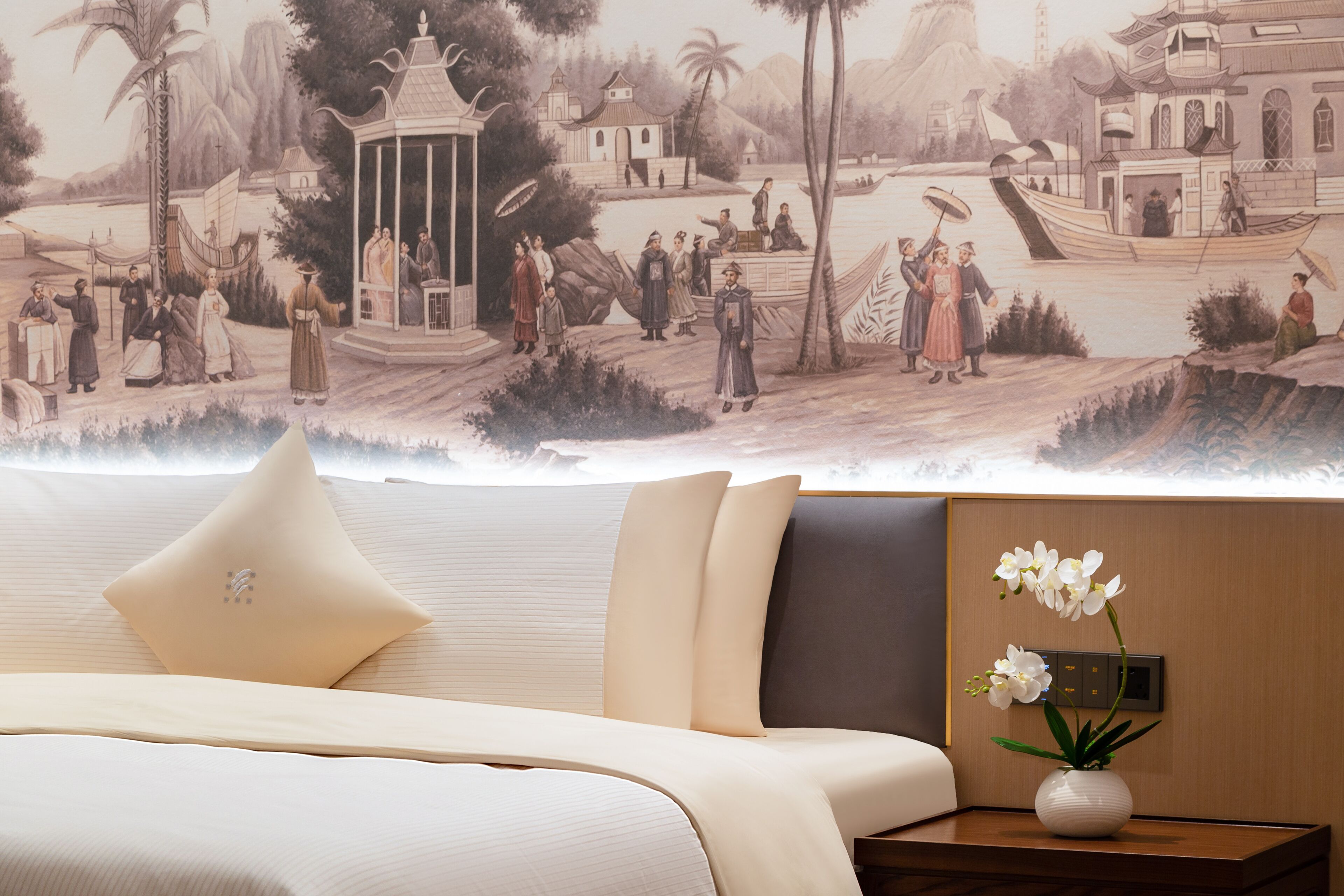 Quarto de hotel com um mural retratando uma cena histórica, uma cama com travesseiro e uma mesinha de cabeceira com um vaso de flores.