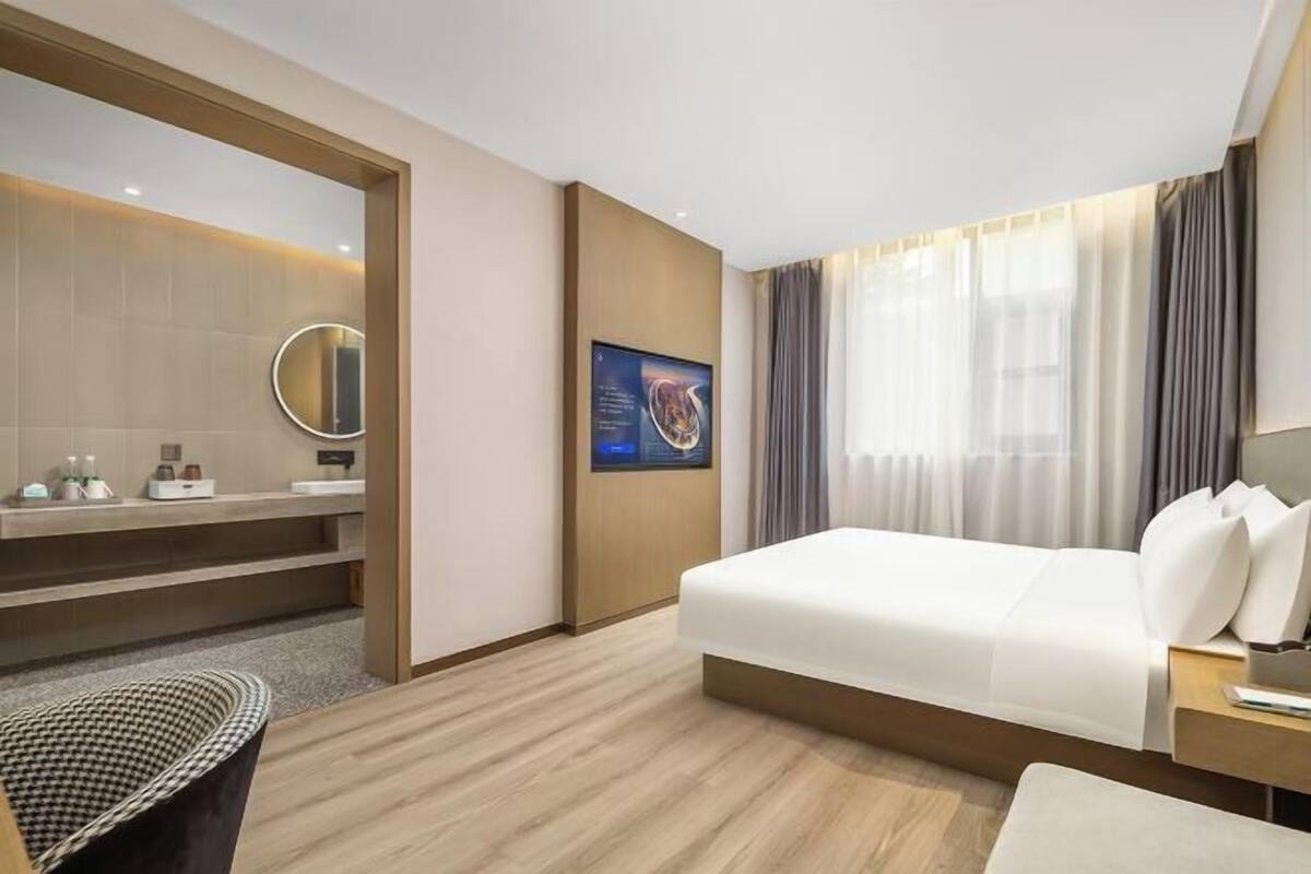Quarto de hotel moderno com uma cama grande, mesa de cabeceira, espelho e vista da cidade através de amplas janelas.