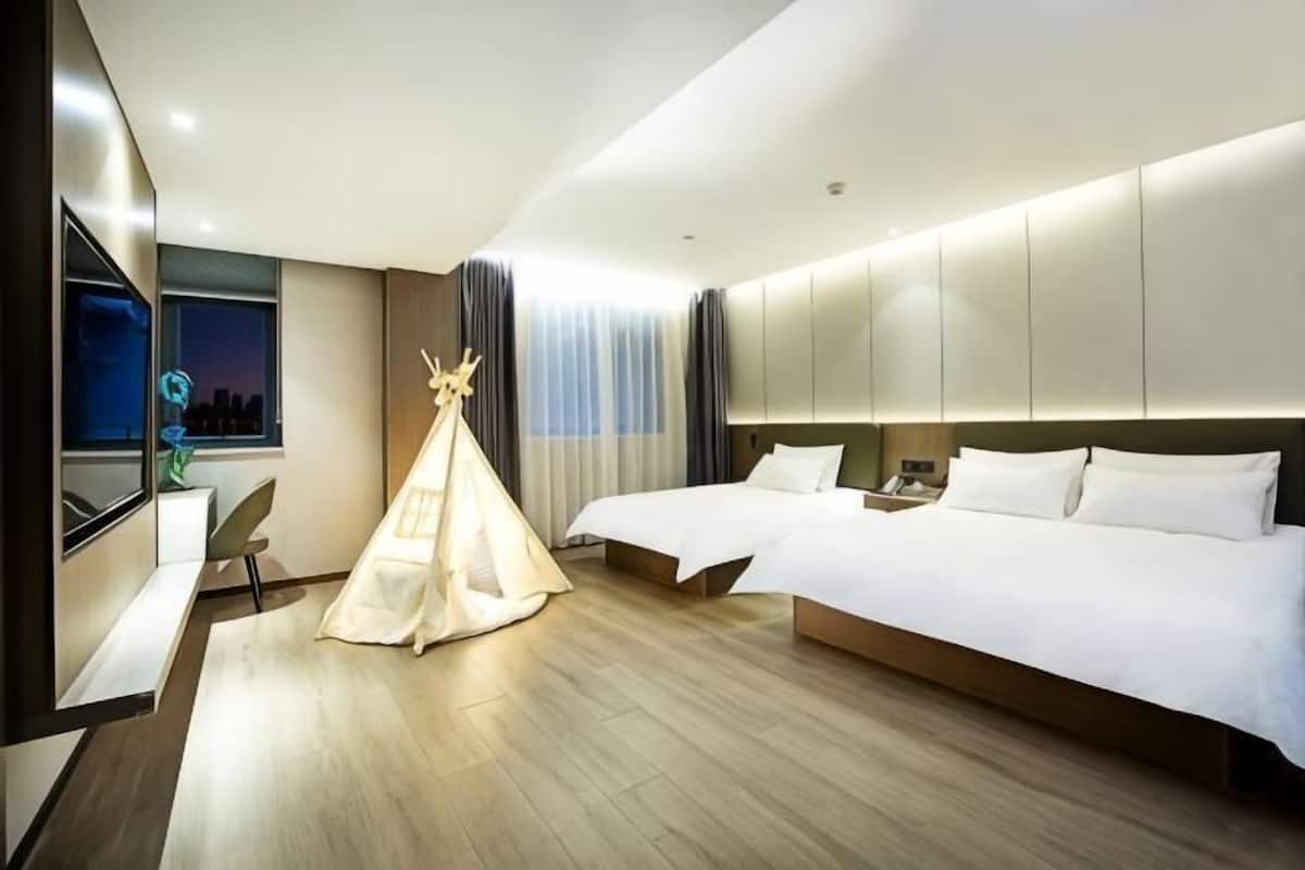 Quarto de hotel moderno com duas camas, uma escrivaninha, uma cadeira e uma janela grande com cortinas.
