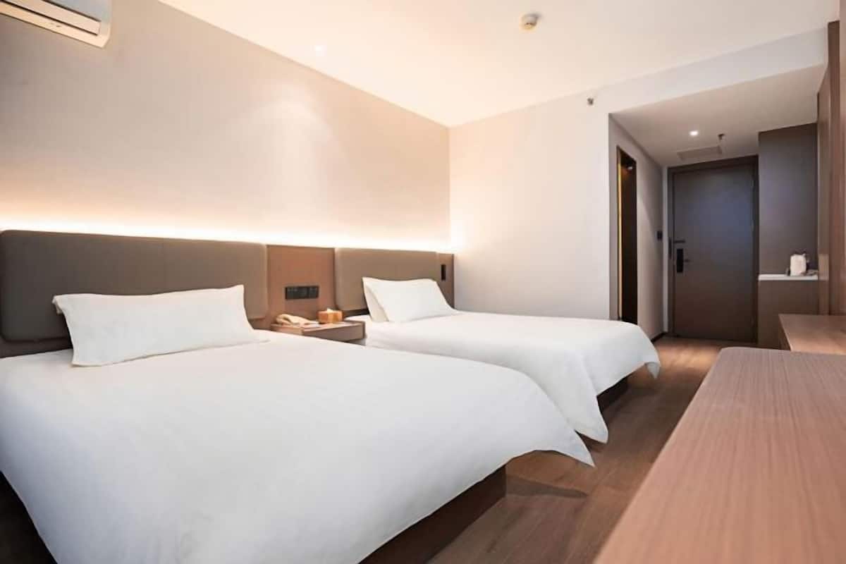 Quarto de hotel com duas camas, uma mesa de cabeceira com abajur e uma escrivaninha de madeira.