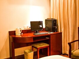 Quarto de hotel com escrivaninha de madeira, computador, cadeira, banquinho pequeno e lixeira.