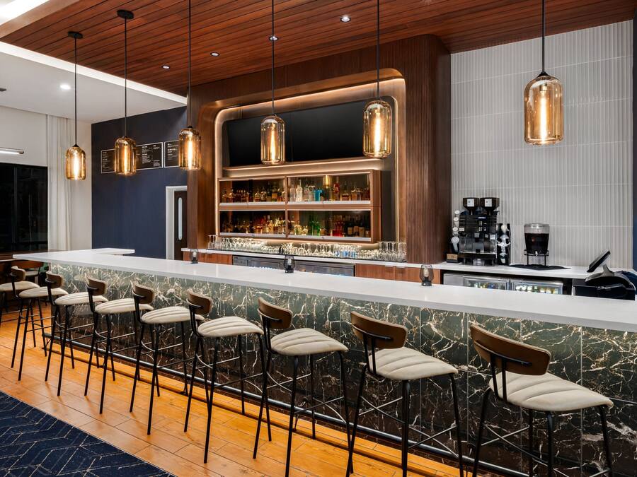 Uma área de bar com balcão de mármore, vários bancos, luminárias pendentes e uma máquina de café.