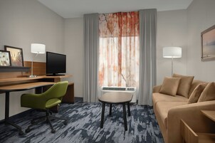 Quarto de hotel com escrivaninha, uma cadeira verde, um sofá marrom, televisão e uma janela com cortinas.