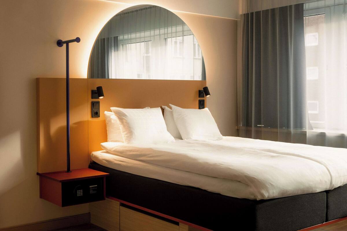 Quarto de hotel moderno com uma cama grande, uma mesa de cabeceira com abajur e uma janela ampla com cortinas.