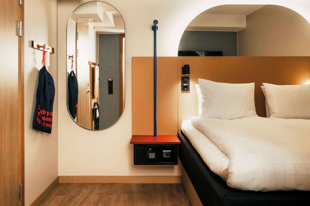 Quarto de hotel com uma cama, um espelho, um porta-casacos e um painel de controlo fixado na parede.