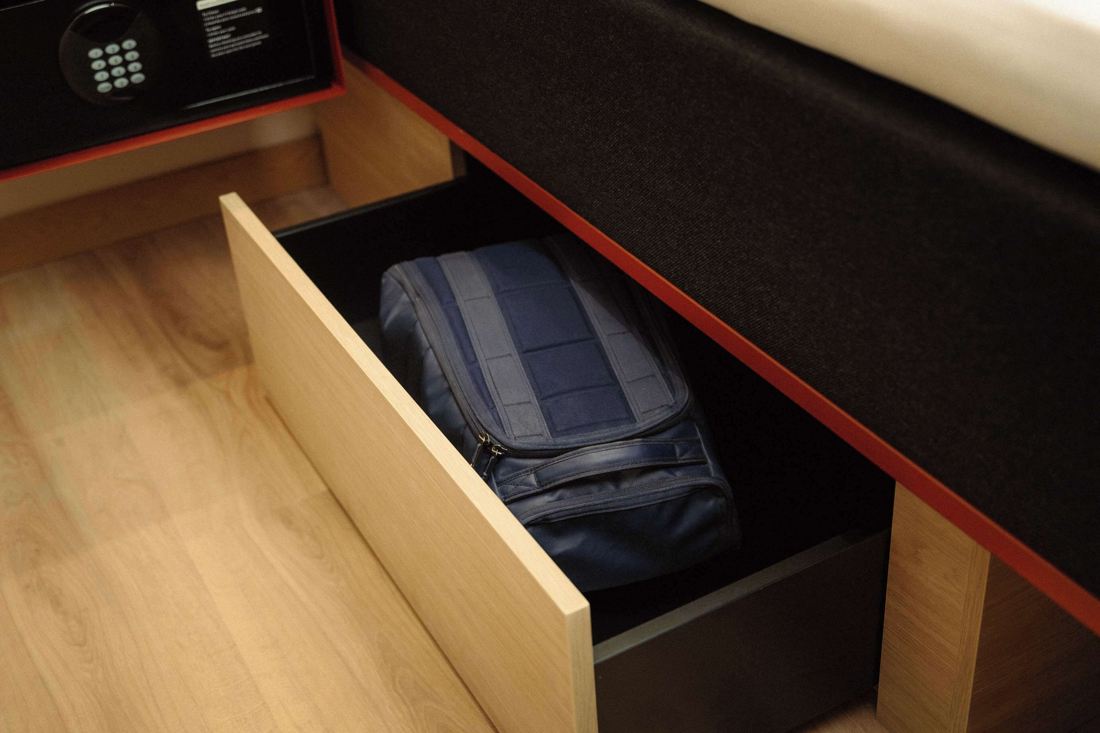 Quarto de hotel com piso de madeira, estrutura de cama preta e uma gaveta com uma bolsa azul.