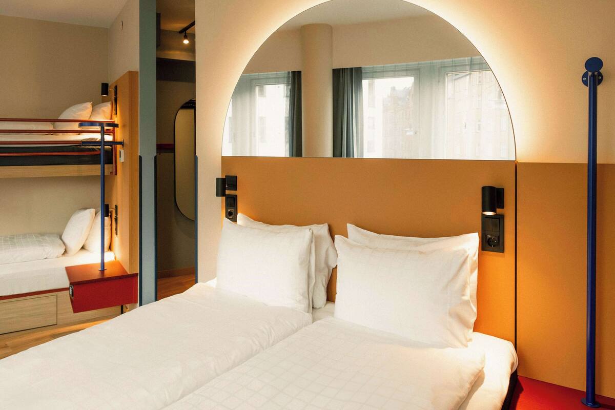 Quarto de hotel com uma cama beliche, uma cama grande com lençóis brancos, um espelho e um abajur azul.