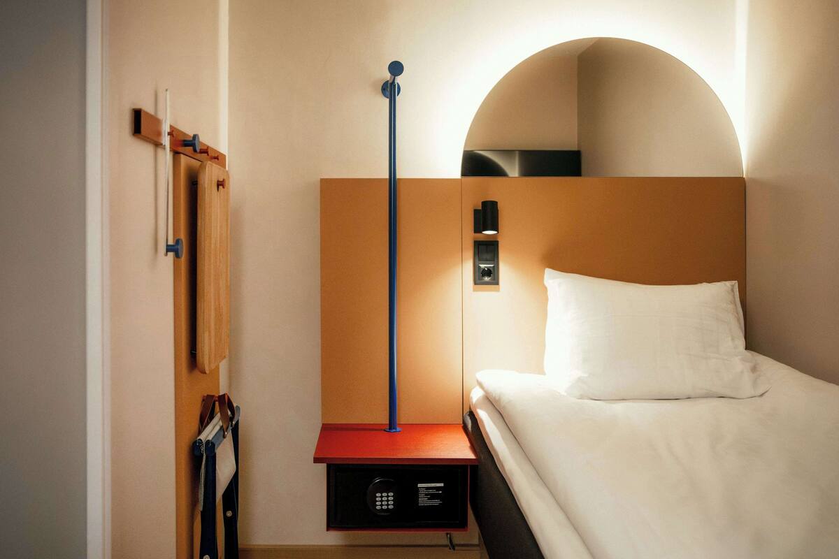 Quarto de hotel com cama, mesa de cabeceira, abajur e painel de controle fixado na parede.