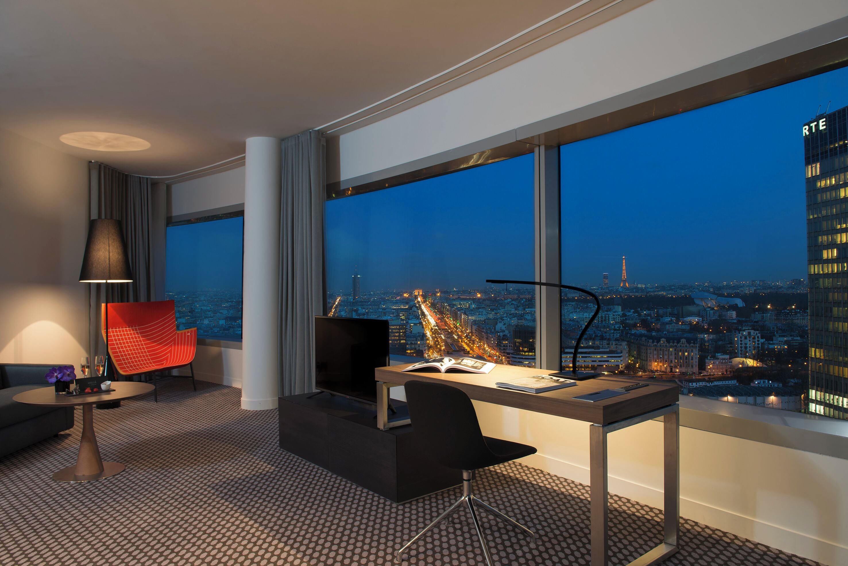 Quarto de hotel moderno com uma janela grande, uma escrivaninha, uma cadeira, uma televisão e um abajur.