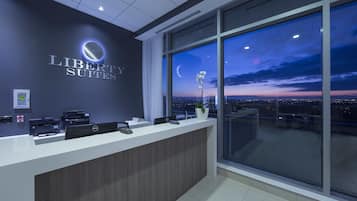 Um balcão de recepção moderno com vista para a cidade ao entardecer, uma placa com a inscrição "Liberty Suites" e uma orquídea em vaso no balcão.