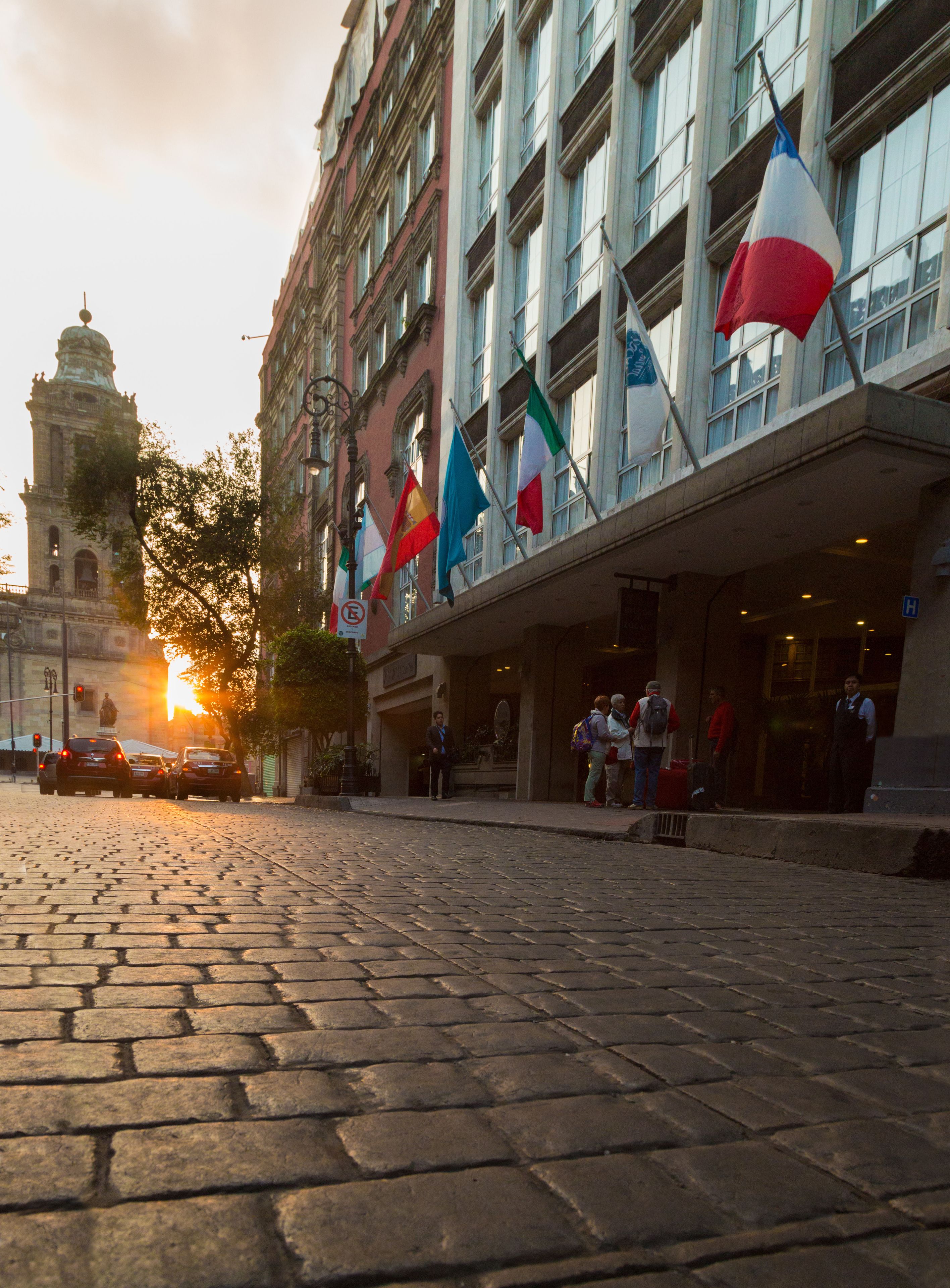 Uma rua de paralelepípedos com um edifício com várias bandeiras, uma estátua e céu claro.