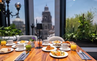 Mesa de jantar com itens de café da manhã e vista para a cidade através de grandes janelas.