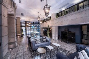 Um hall amplo com teto alto, área de estar com sofá e poltronas, uma vitrine de vidro com objetos decorativos e uma grande janela com vista para o céu.