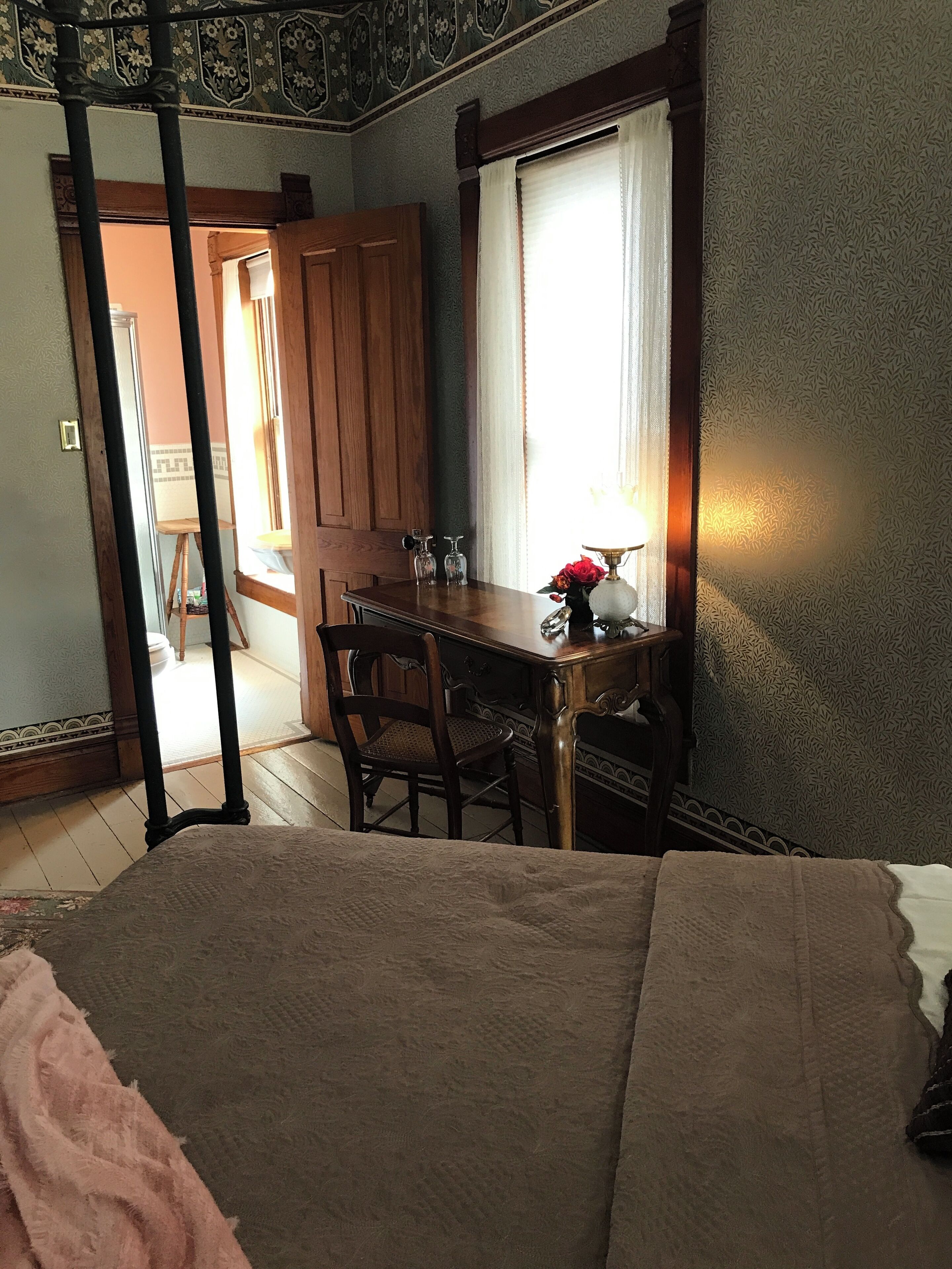 Quarto com cama, escrivaninha de madeira, cadeira e janela com cortinas.