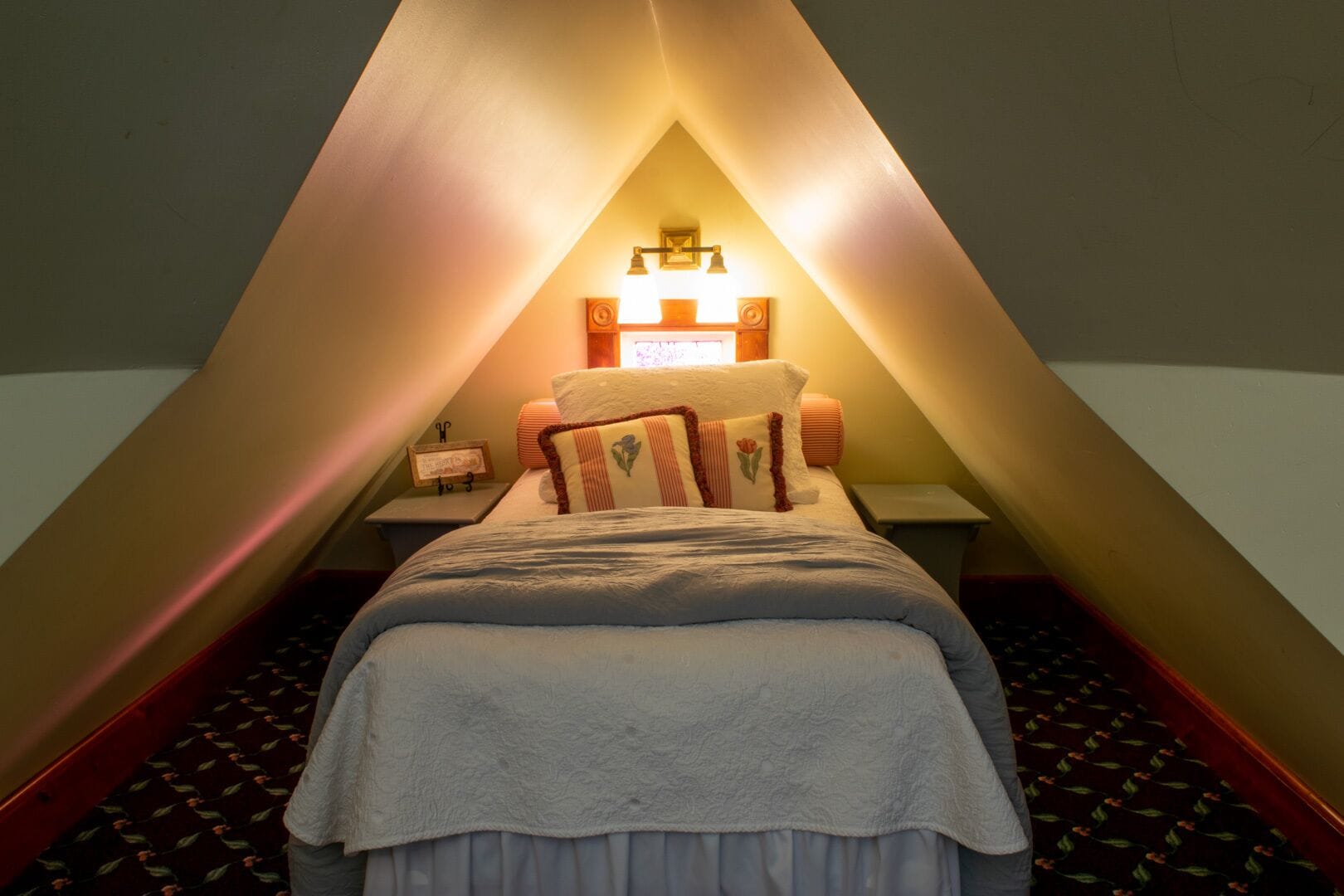 Quarto sótão acolhedor em formato triangular, com cama, almofadas e mesa de cabeceira.