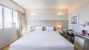 Quarto de hotel com uma cama grande, escrivaninha, cadeira, mini-frigorífico e vista para a cidade.