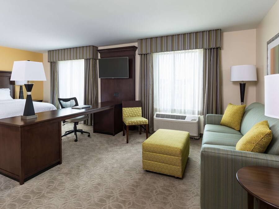 Quarto de hotel com cama, escrivaninha, duas cadeiras, sofá e televisão.
