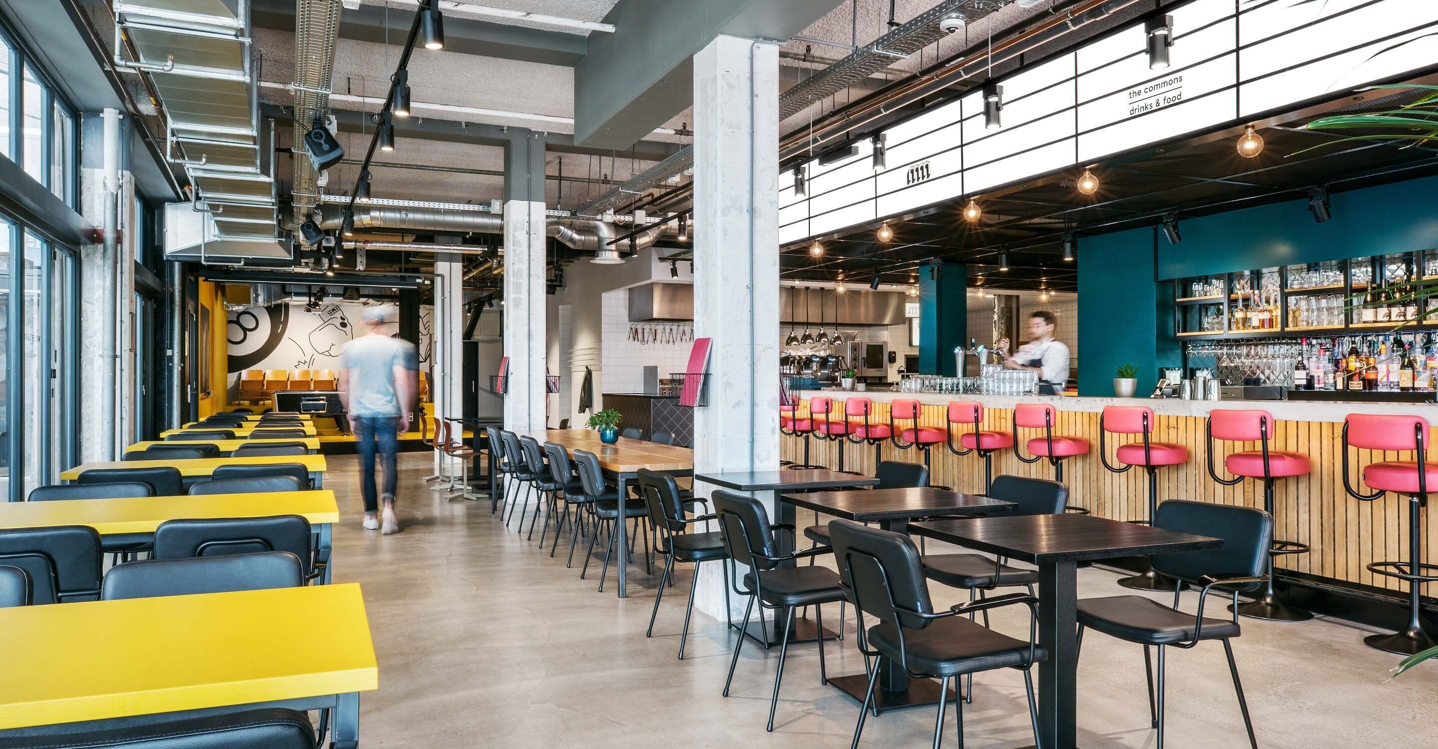 Um restaurante moderno com mesas amarelas, cadeiras pretas e um espaço de bar com banquetas vermelhas.