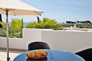 Uma mesa redonda com uma tigela de frutas, uma cadeira de vime preta e um guarda-sol bege numa varanda com vista para o mar.