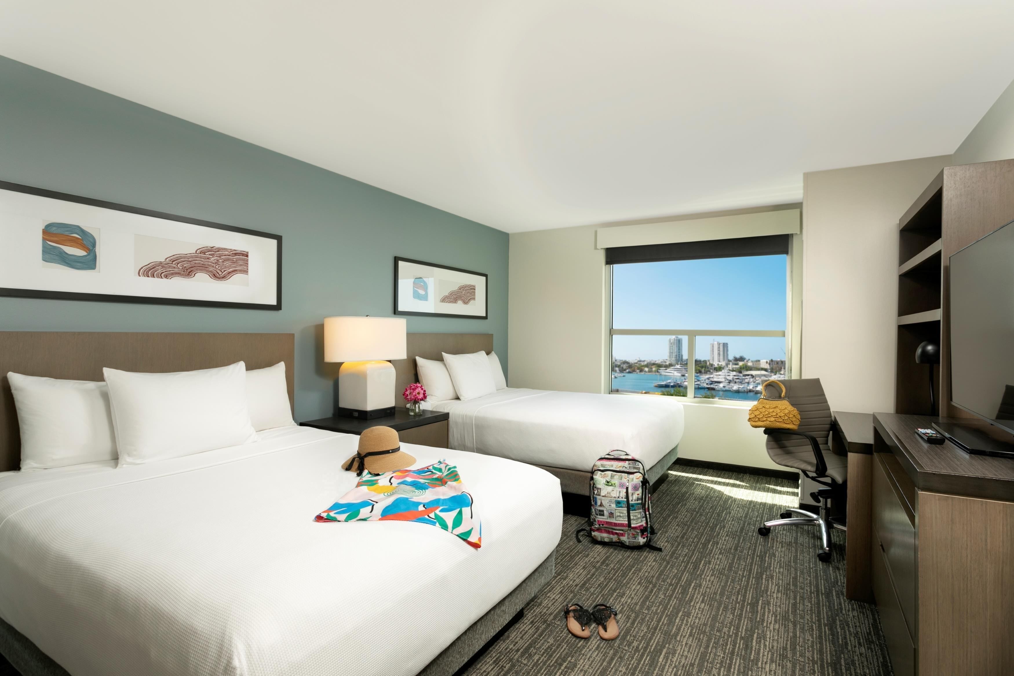 Quarto de hotel com duas camas, uma escrivaninha, uma cadeira, uma televisão e uma janela grande com vista para a cidade.
