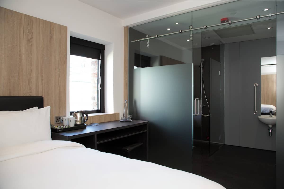 Quarto de hotel moderno com cama, escrivaninha com cafeteira, box de vidro e vista para a cidade.
