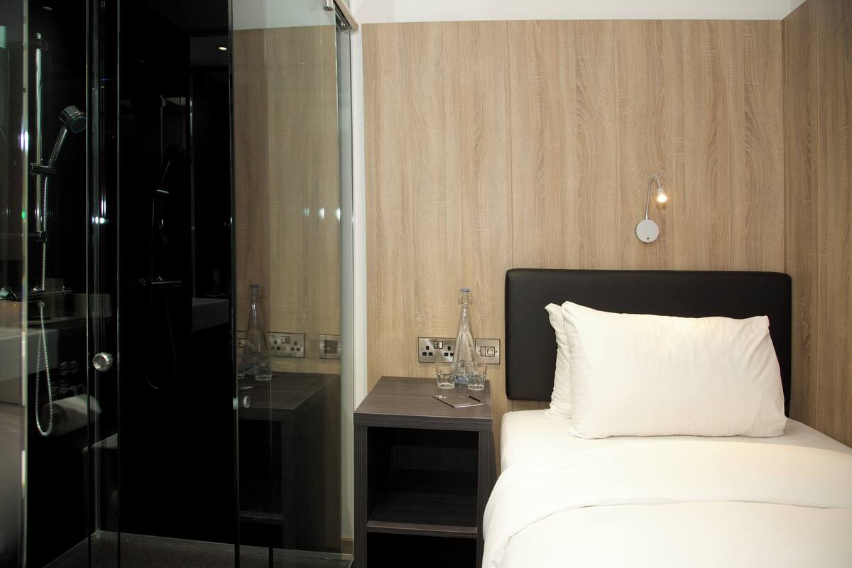 Quarto de hotel com chuveiro em cabine de vidro, mesa de cabeceira com garrafa e copos, e cama com travesseiro branco.