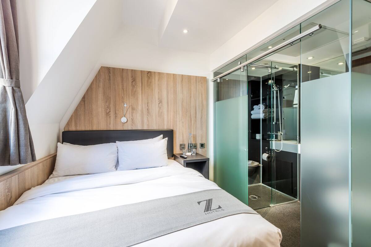 Quarto de hotel moderno com cabeceira de madeira, cama com roupa de cama branca, chuveiro com divisória de vidro e um porta-toalhas visível.