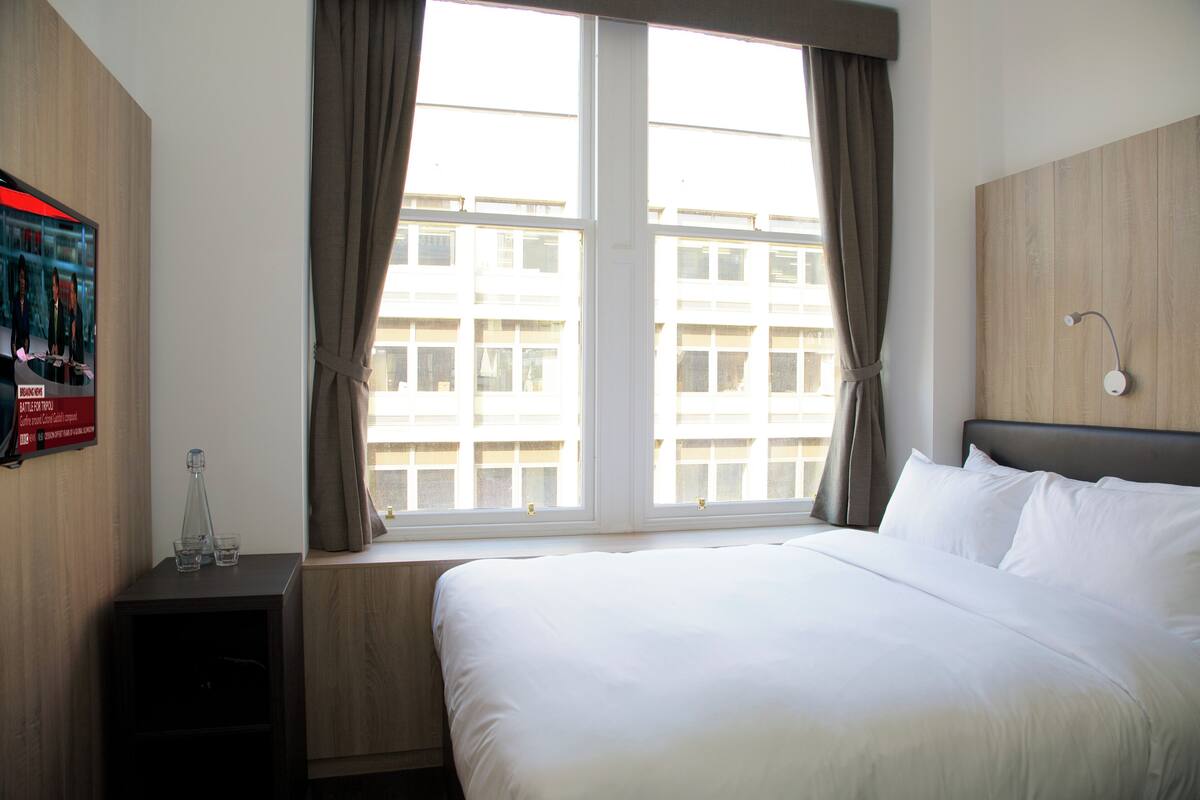 Quarto de hotel com uma cama grande, uma janela com cortinas, mesa de cabeceira com garrafa e copos, e televisão fixada na parede.