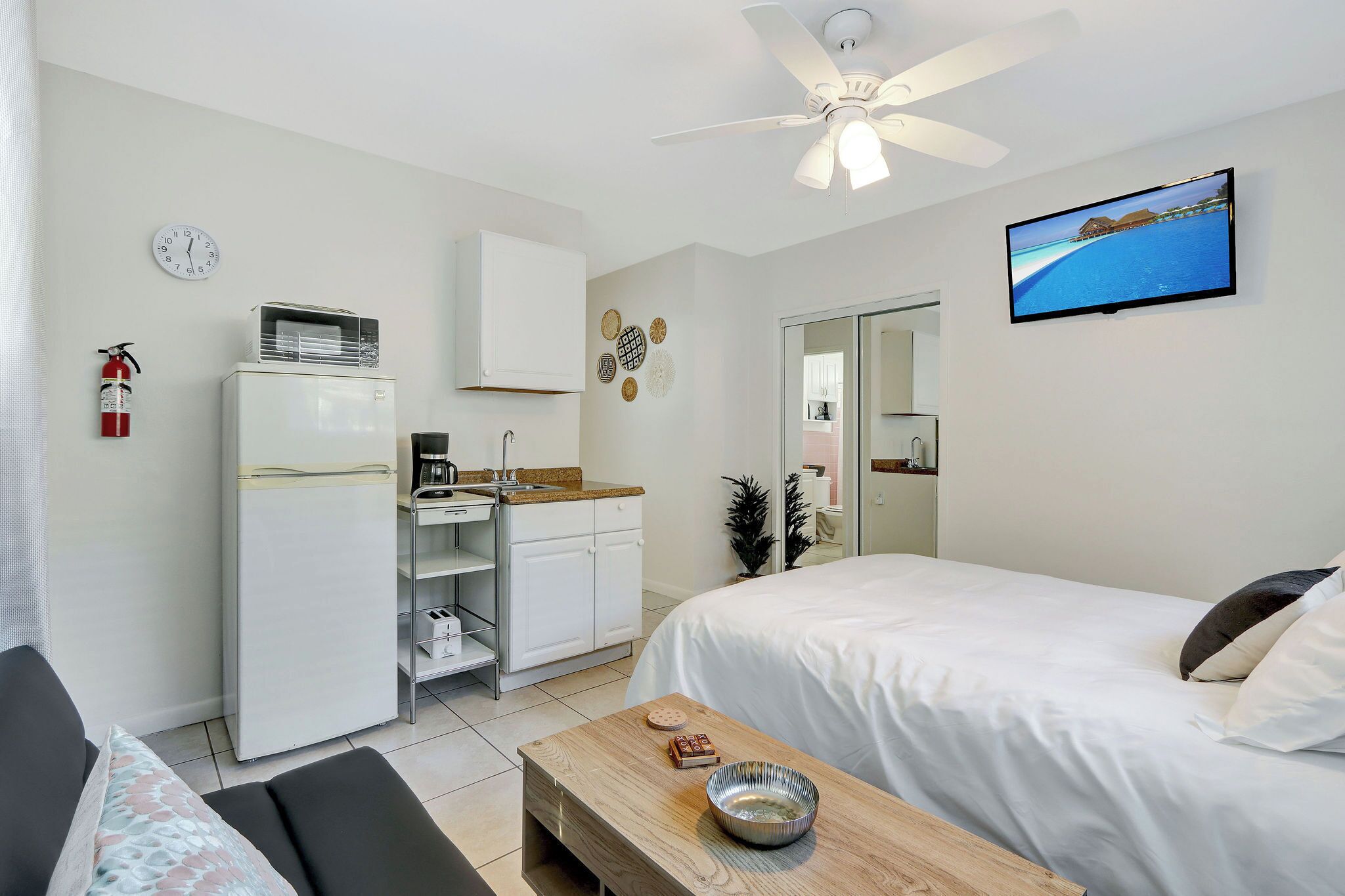 Quarto de hotel moderno com cama, escrivaninha, frigorífico, máquina de café, televisão e ventoinha de teto.