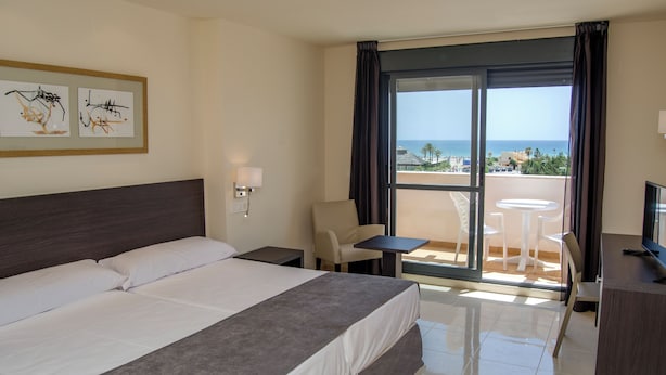 Quarto de hotel com uma cama grande, uma escrivaninha, uma cadeira, uma TV e uma varanda com vista para o mar.