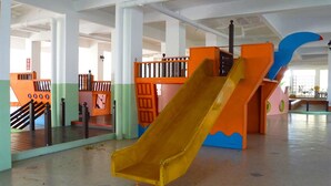 Uma área de recreação com um escorrega amarelo, estruturas de jogo em laranja e azul, e uma escada.
