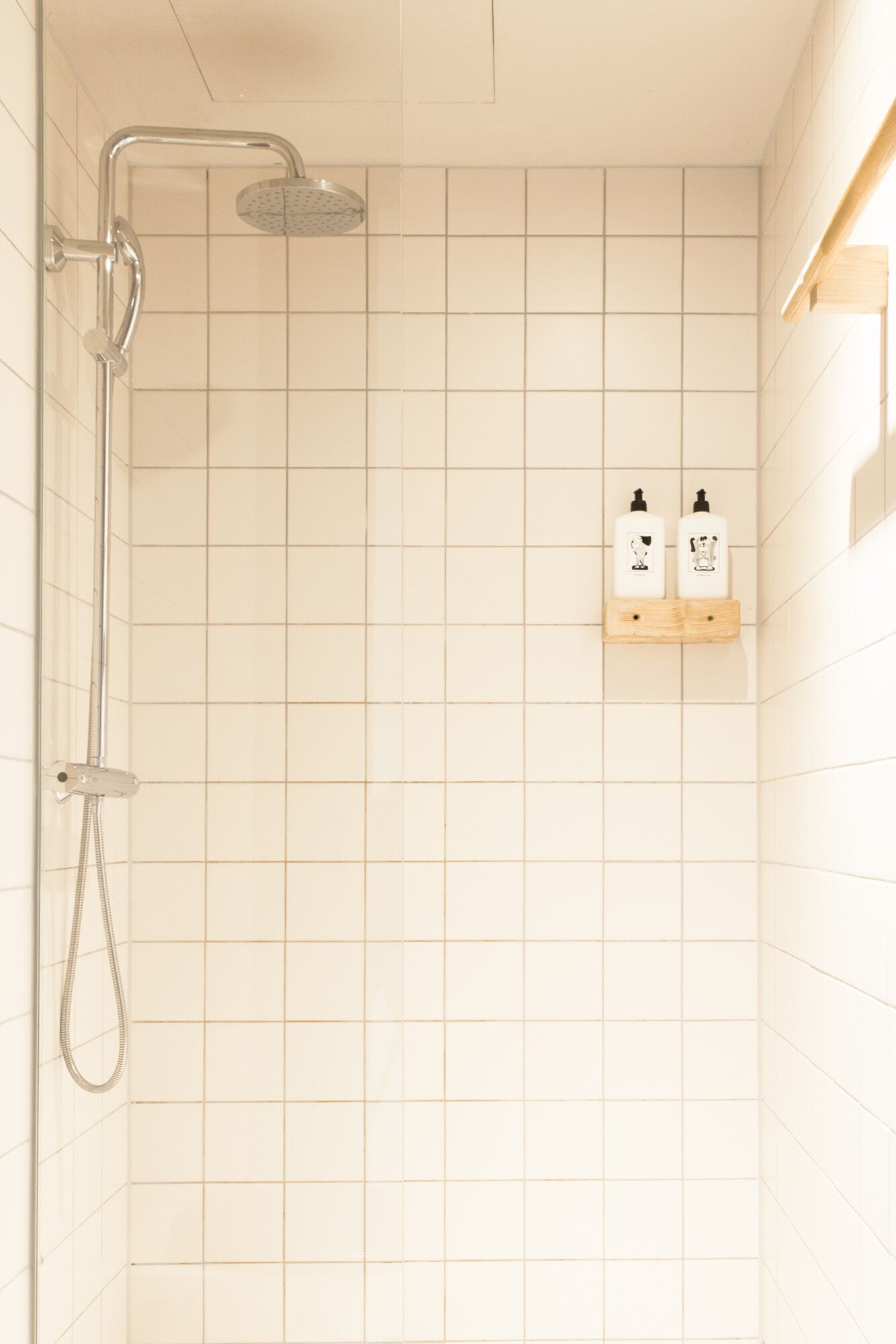 Um chuveiro com chuveiro manual e ducha de teto, paredes azulejadas e dois dispensadores de sabonete em uma prateleira de madeira.