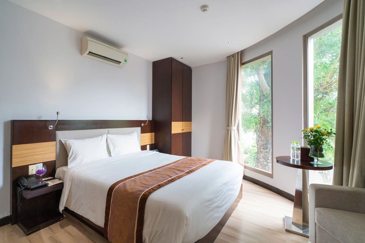 Quarto de hotel com uma cama grande, mesa de cabeceira com telefone e copo, pequena mesa com um vaso de flores e janela com vista para árvores.