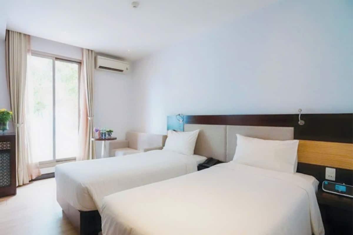 Quarto de hotel com duas camas, mesa de cabeceira, cortinas na janela e um aparelho de ar condicionado.