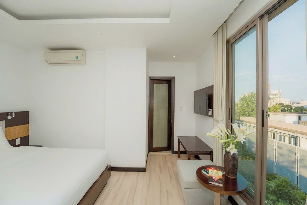 Quarto de hotel moderno com cama, uma pequena mesa com um vaso de flores, TV fixada na parede e uma grande janela com vista para a cidade.