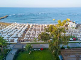 Um hotel à beira-mar com fileiras de espreguiçadeiras e guarda-sóis, um cais que se estende para o mar, e uma placa com a inscrição "CAMARA".