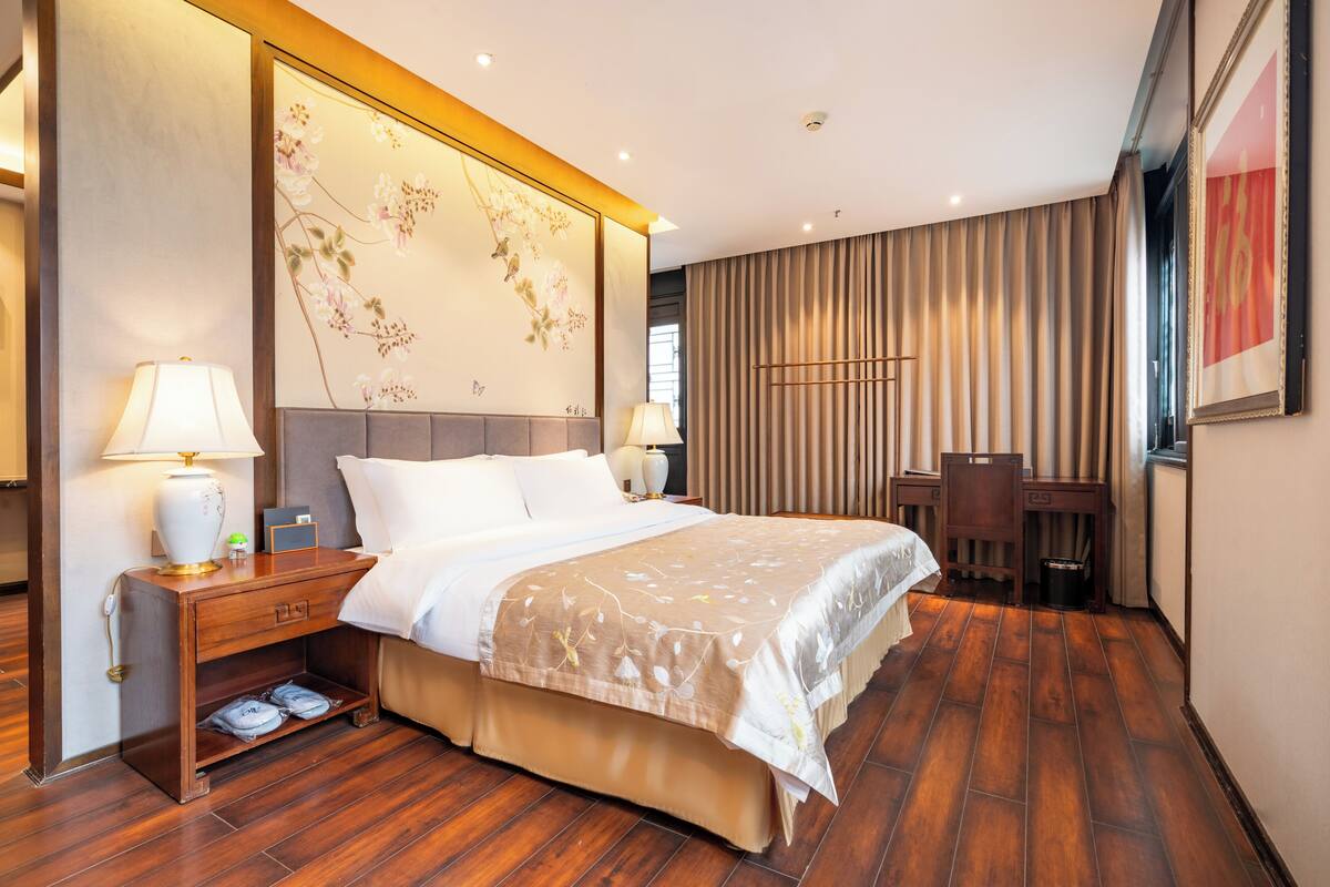 Quarto de hotel com uma cama grande, abajures, mesa de cabeceira de madeira, escrivaninha e uma cadeira.