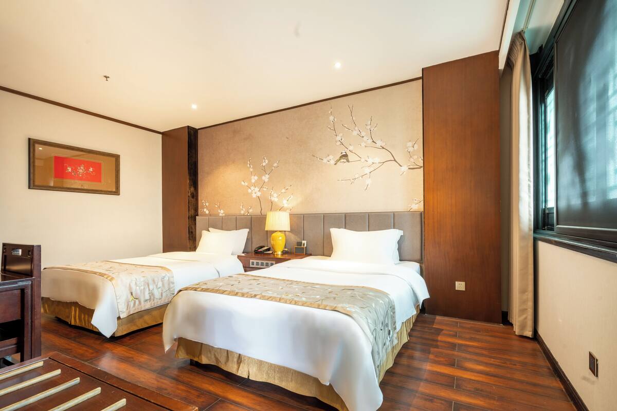 Quarto de hotel com duas camas, piso de madeira, luminária fixada na parede e uma moldura com quadro.